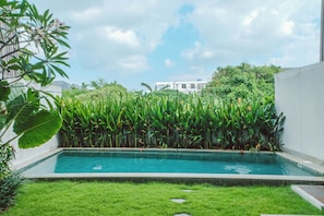 Uma piscina retangular com área gramada na frente, cercada por vegetação densa e um edifício ao fundo.
