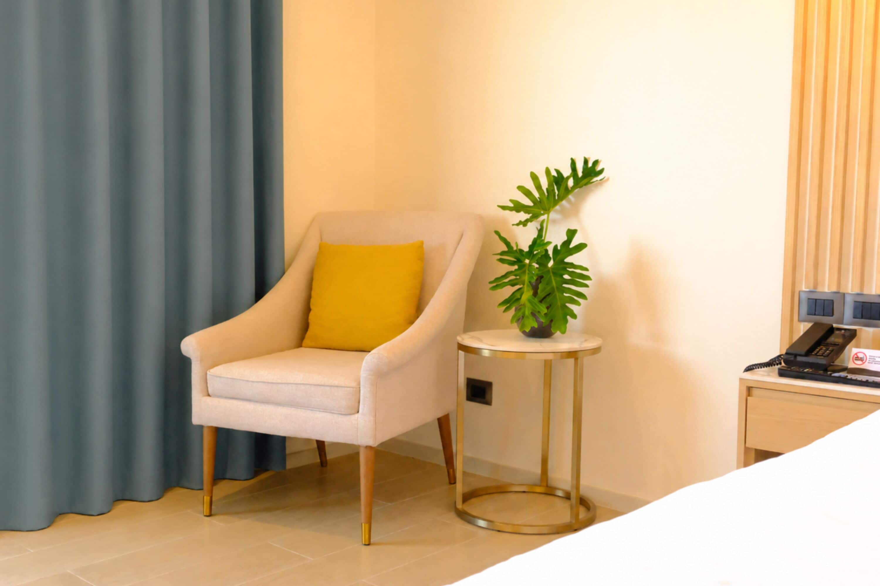 Quarto de hotel com uma poltrona de cor clara com almofada amarela, uma mesinha redonda com uma planta, mesa de cabeceira com telefone e cortina.