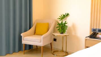 Quarto de hotel com uma poltrona de cor clara com almofada amarela, uma mesinha redonda com uma planta, mesa de cabeceira com telefone e cortina.