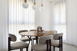 Uma sala de jantar com uma mesa de madeira redonda, seis cadeiras estofadas e três luminárias pendentes.