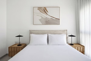 Uma cama bem arrumada com duas mesas de cabeceira, cada uma com uma luminária, em um quarto simples com uma parede lisa e uma única obra de arte.