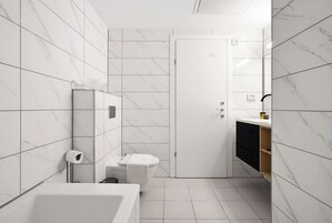 Um banheiro moderno com azulejos de mármore branco, uma banheira branca, vaso sanitário e um espelho sobre um móvel de madeira.