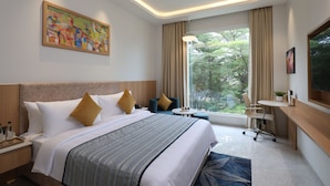 Quarto de hotel com uma cama grande, uma escrivaninha, uma cadeira, uma janela com vista para árvores e um quadro na parede.