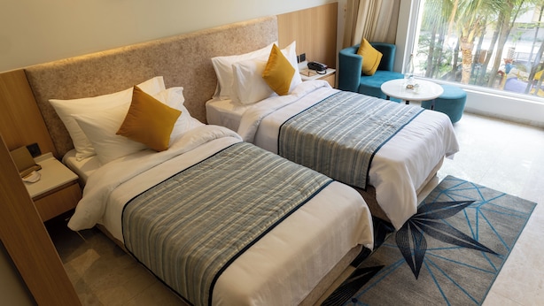Quarto de hotel com duas camas, uma mesinha e vista para palmeiras.