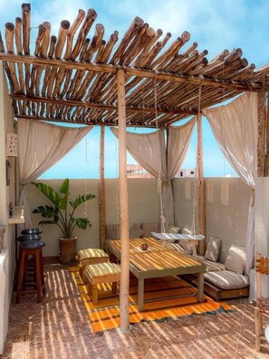 Uma área externa com mesa e bancos de madeira, uma cadeira suspensa e um vaso com planta.