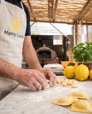 Uma pessoa preparando comida, especificamente massa, em uma cozinha com forno de tijolos e limões frescos ao fundo.