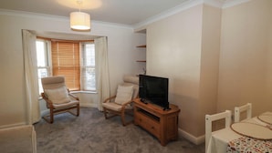 Uma sala de estar com dois poltronas, uma televisão sobre um suporte de madeira, uma mesa de jantar com toalha de mesa estampada e uma luminária no teto.