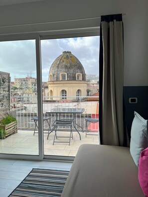 Quarto de hotel com varanda, cama, sofá e vista para um edifício histórico.