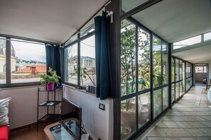 Quarto iluminado pelo sol, com janelas amplas, uma pequena mesa com uma planta e vista para edifícios.