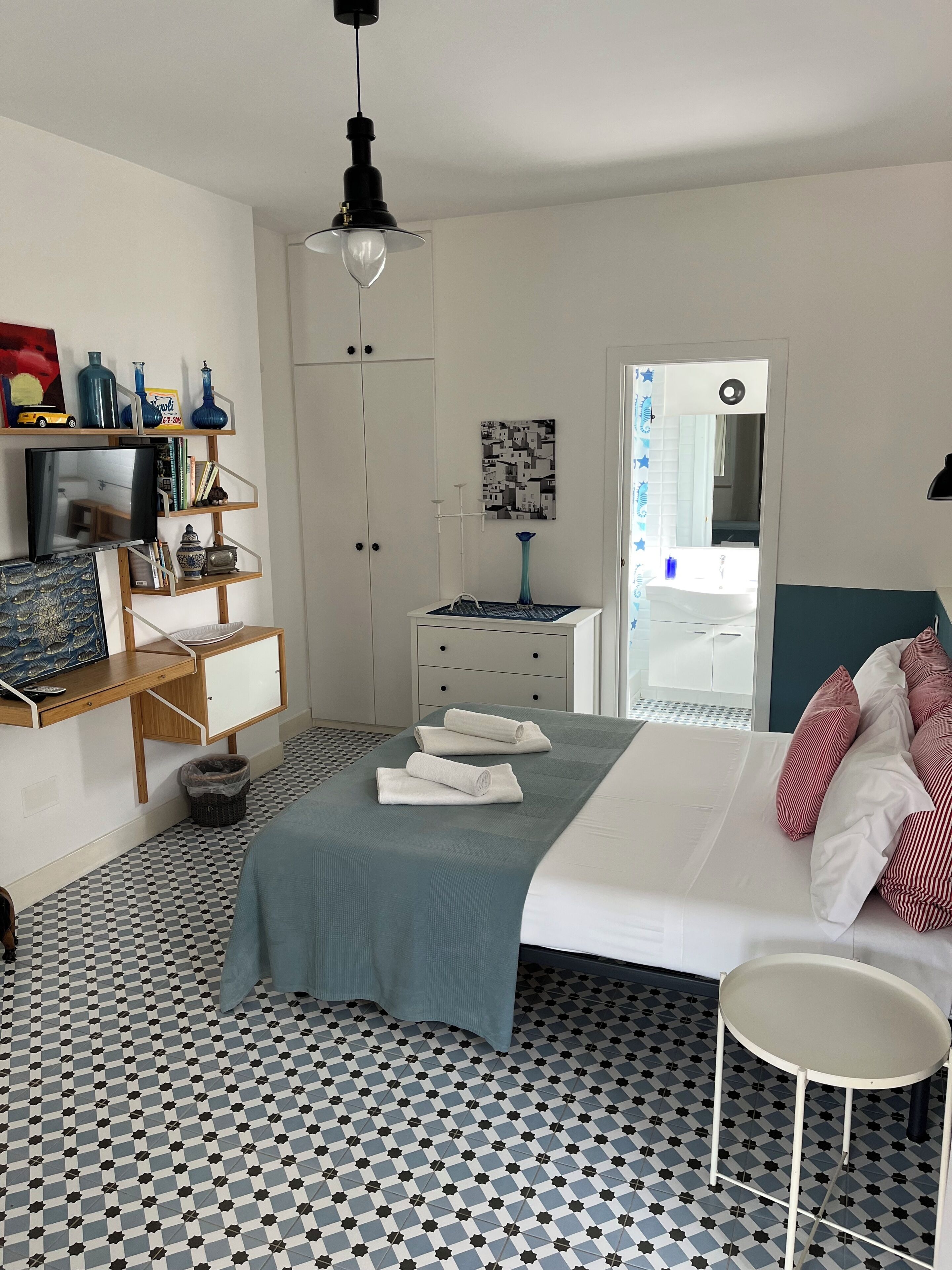 Quarto com cama, mesa de cabeceira, poltrona, guarda-roupa e prateleira com objetos decorativos.