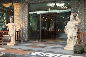 Entrada de um edifício com pilares de pedra, uma estátua de uma figura segurando um cajado e uma placa com a inscrição 'RESTAURANT'.