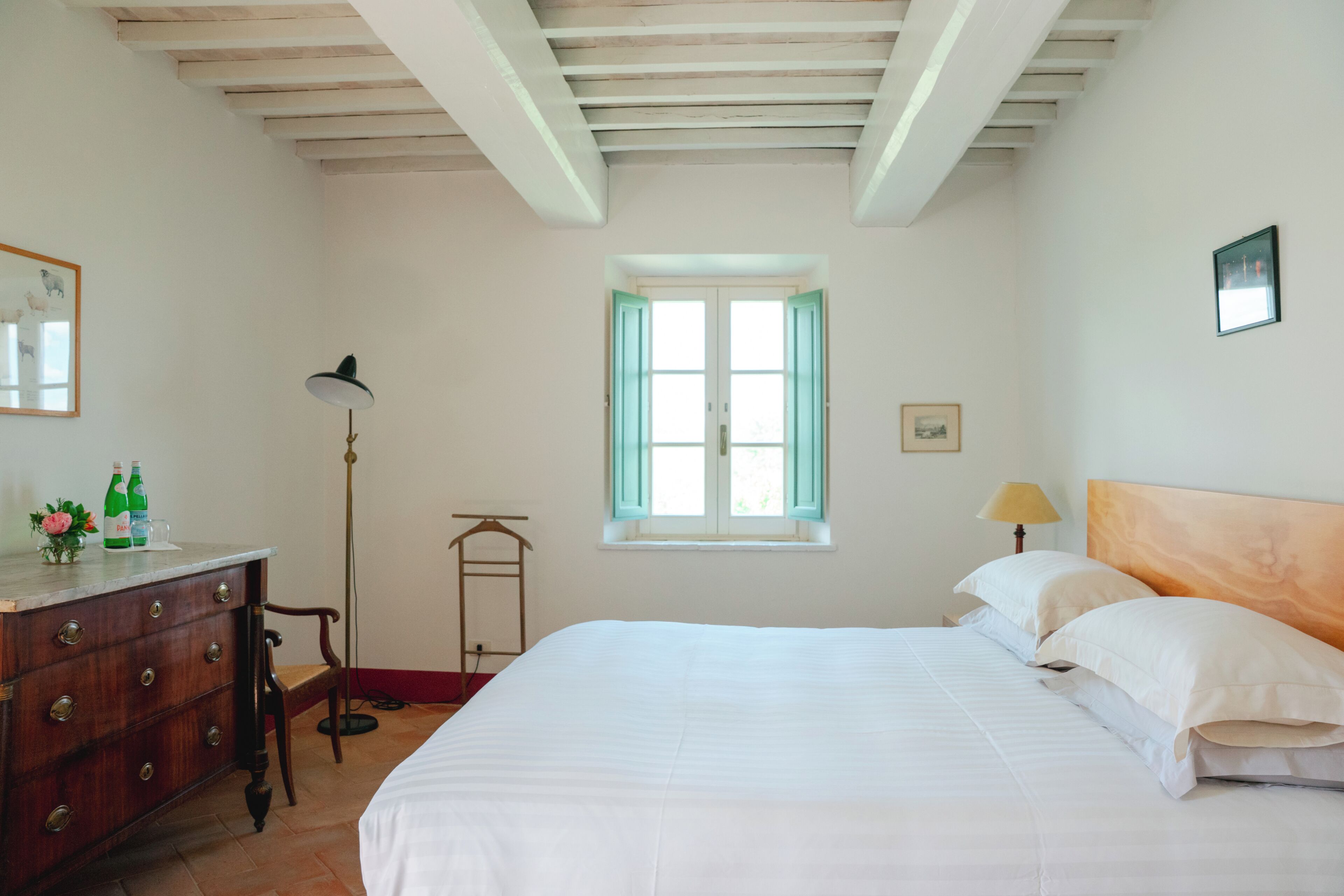 Um quarto com uma cama, cômoda, luminária, janela e um quadro na parede.