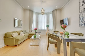 Uma sala de estar moderna com sofá, mesa de centro de vidro, mesa de jantar com cadeiras, televisão e uma janela grande com cortinas.