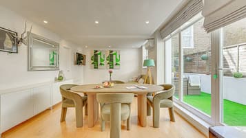 Uma área de refeições moderna com uma mesa redonda, seis cadeiras e uma janela grande com vista para uma parede de tijolos e vegetação lá fora.