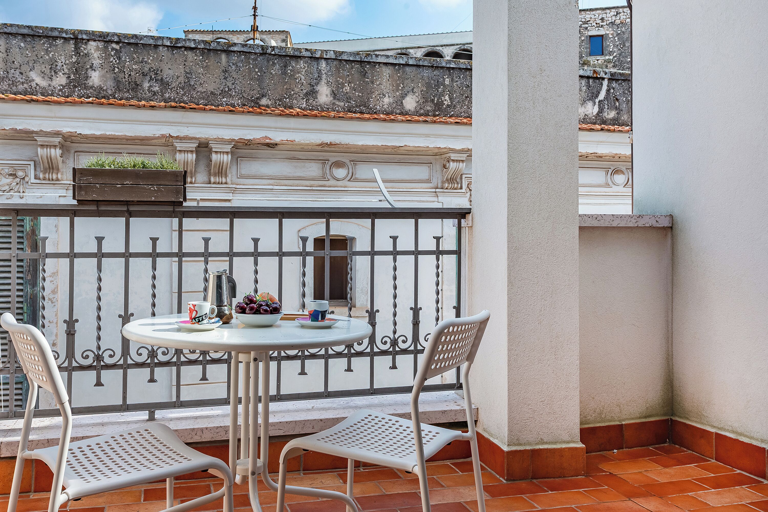 Varanda com mesa branca e duas cadeiras, um vaso pequeno com planta e vista para um edifício com telhado de azulejos.