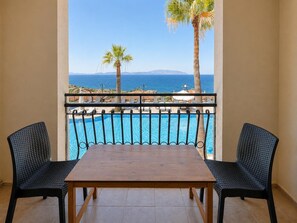 Varanda com mesa de madeira e duas cadeiras, com vista para uma piscina e para o mar, com palmeiras.