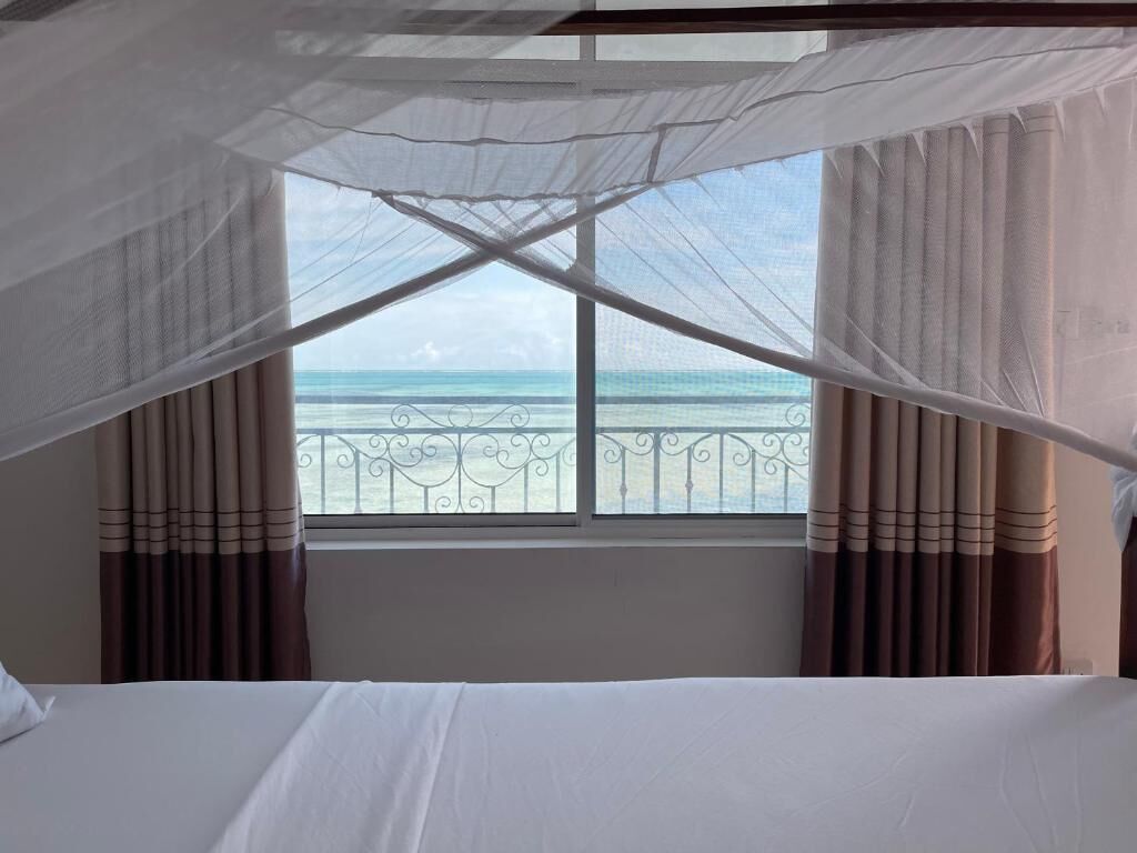 Um quarto com cama, janela com cortinas e vista para o oceano.