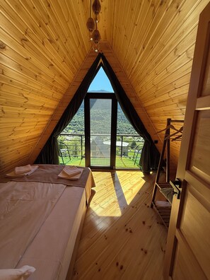 Quarto de sótão com piso de madeira, janela grande, cama e escada que dá para uma varanda.