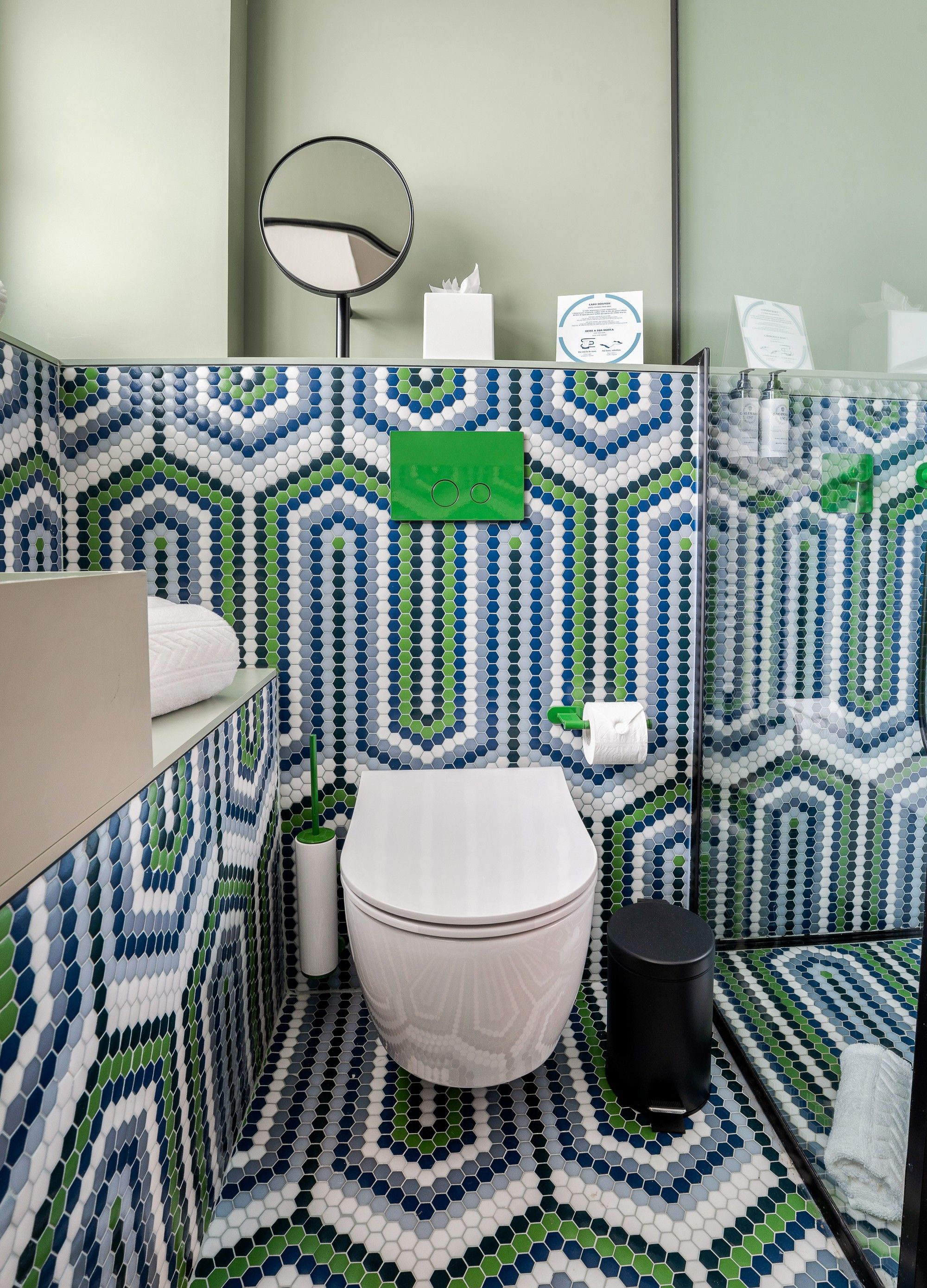 Casa de banho com sanita branca, dispensador de sabonete verde e parede com azulejos estampados.