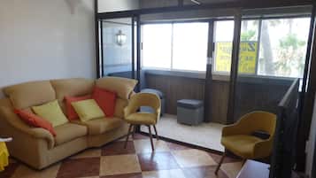 Um quarto com um sofá bege, duas poltronas, uma mesa pequena e uma janela com vista para palmeiras.