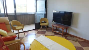 Quarto com televisão, duas cadeiras amarelas, sofá com almofadas laranja e chão xadrez.