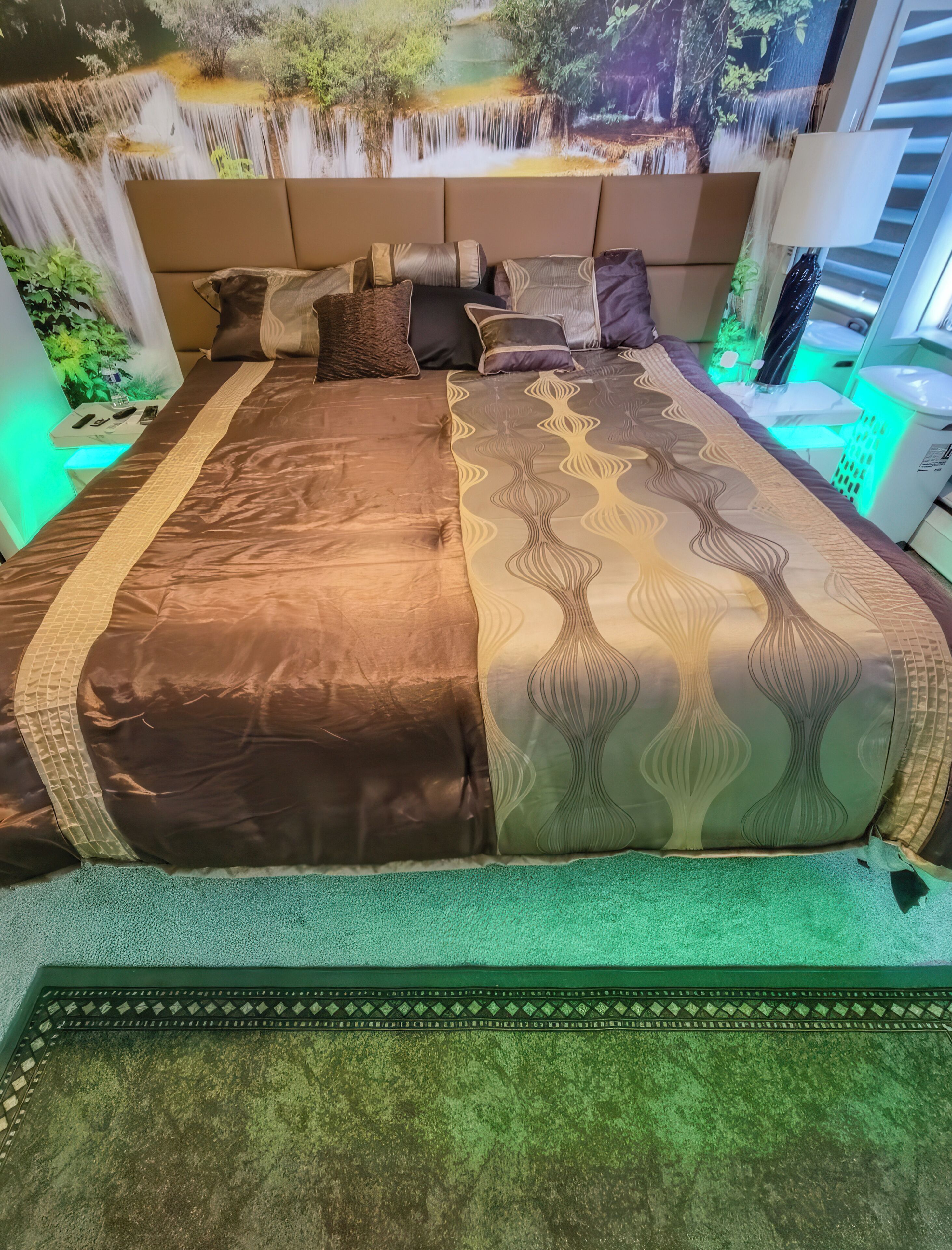 Uma cama com colcha estampada, várias almofadas, uma cabeceira e uma mesa de cabeceira com abajur.