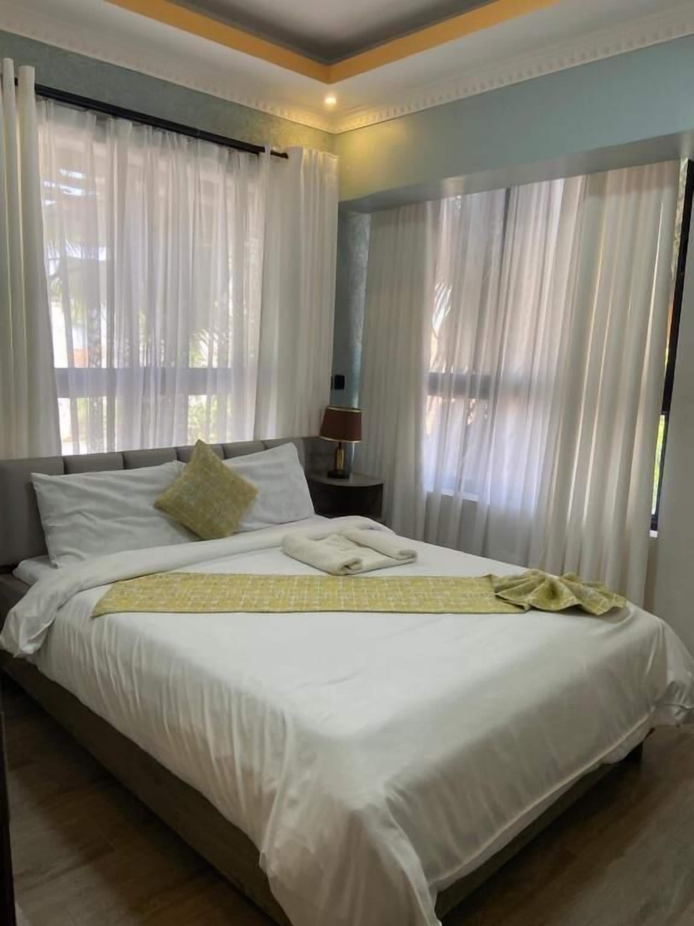 Cama bem arrumada com lençóis brancos, um cobertor amarelo e um travesseiro. Tem uma mesa de cabeceira com uma luminária ao lado da cama.