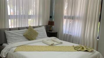 Cama bem arrumada com lençóis brancos, um cobertor amarelo e um travesseiro. Tem uma mesa de cabeceira com uma luminária ao lado da cama.