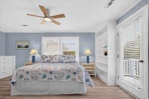 Quarto com uma cama, duas mesas de cabeceira com luminárias, um ventilador de teto, uma janela com persianas e uma estante de livros.