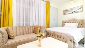 Quarto de hotel com sofá bege, mesa de centro branca, cama com cabeceira estofada e cortinas amarelas.
