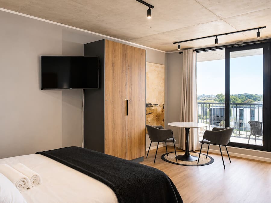 Quarto de hotel moderno com cama, televisão de tela plana, armário de madeira, varanda com mesa e cadeiras, e uma grande janela com vista.