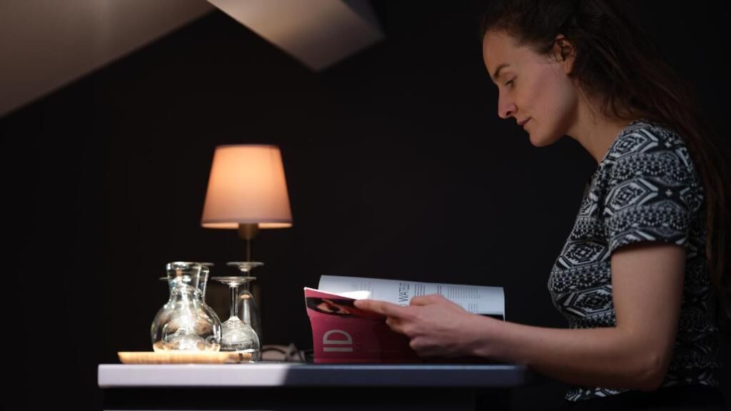 Uma pessoa lendo um livro numa mesa com uma lâmpada e duas garrafas de vidro.