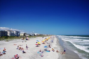 Uma praia com pessoas se banhando ao sol e nadando, guarda-sóis e edifícios ao fundo.