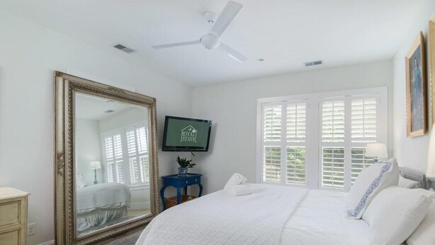 Quarto com um espelho grande, cama com roupa de cama branca, ventilador de teto, televisão e janela com persianas.