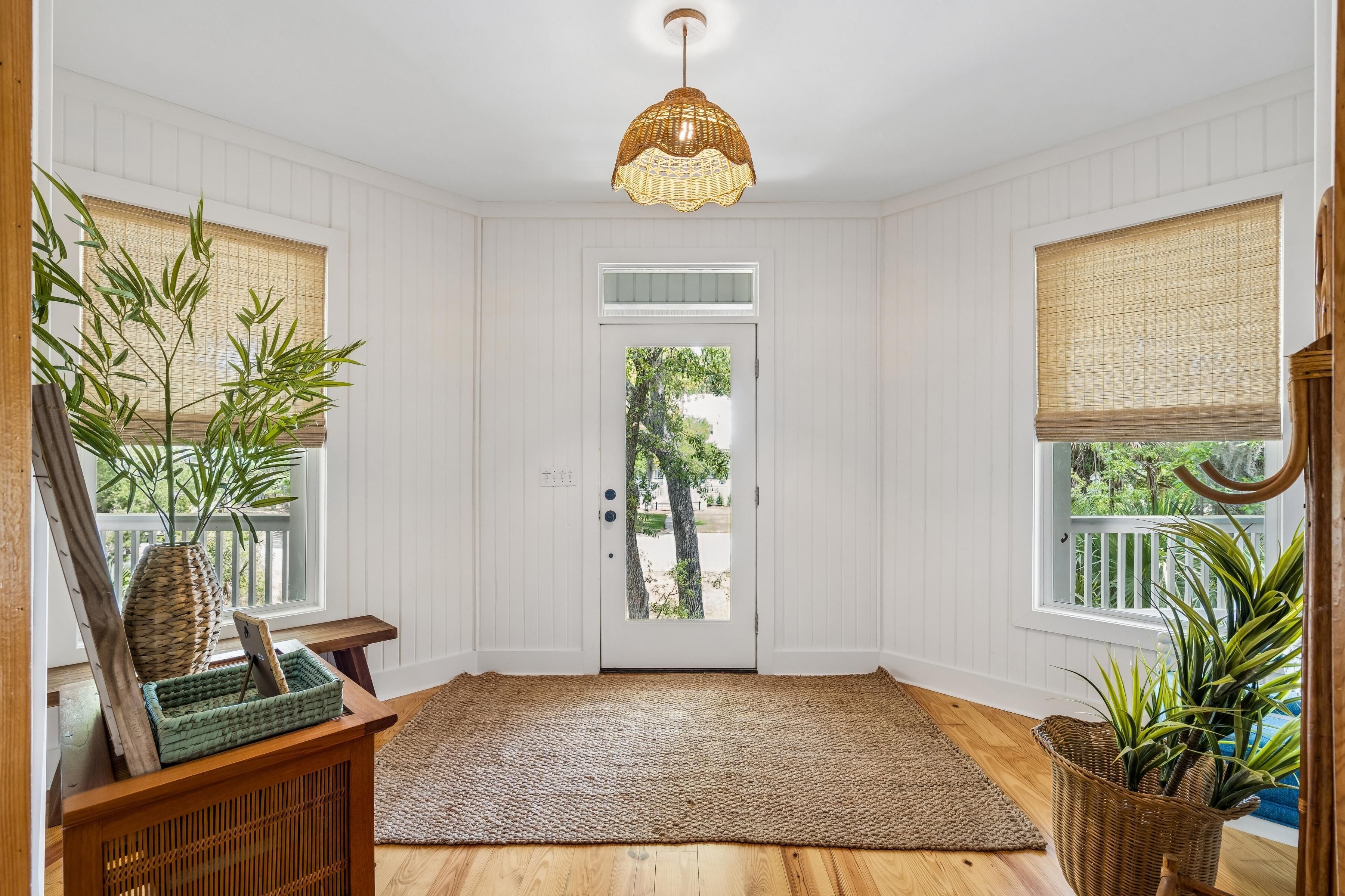 Um quarto com piso de madeira, paredes brancas com painéis, uma porta grande com painel de vidro, uma cesta de vime com plantas e um lustre.