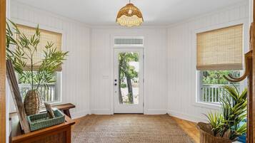 Um quarto com piso de madeira, paredes brancas com painéis, uma porta grande com painel de vidro, uma cesta de vime com plantas e um lustre.