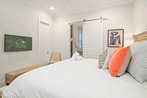 Quarto de hotel com uma cama, um banquinho, uma cadeira, uma porta deslizante e um quadro na parede.