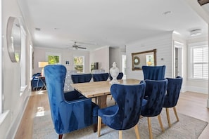 Uma sala de jantar com mesa de madeira e cadeiras estofadas em azul, paredes brancas e ventilador de teto.