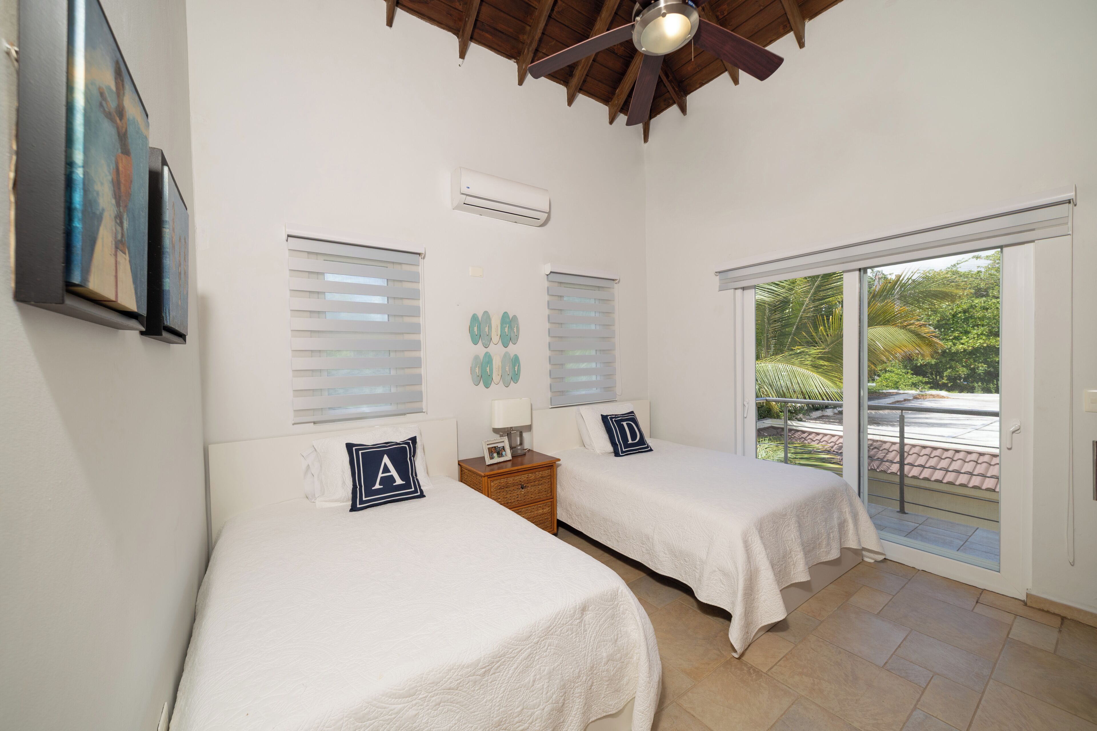 Quarto com duas camas, ventilador de teto, aparelho de ar condicionado, janela com persianas e vista para o verde lá fora.