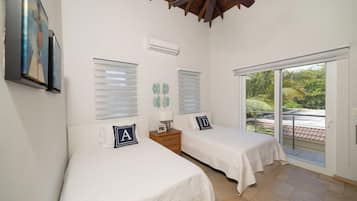 Quarto com duas camas, ventilador de teto, aparelho de ar condicionado, janela com persianas e vista para o verde lá fora.