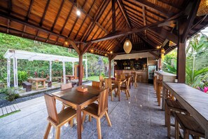 Uma área de refeições ao ar livre coberta, com mesas e cadeiras de madeira, um bar e vista para vegetação exuberante.
