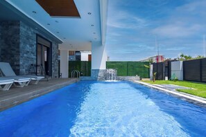 Uma piscina moderna com uma cascata, rodeada por um relvado bem cuidado e um muro de pedra.