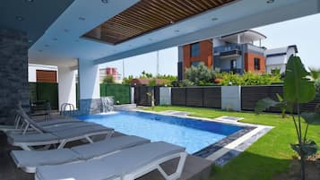 Uma área moderna de piscina ao ar livre com espreguiçadeiras, um muro de pedra e um gramado bem cuidado.