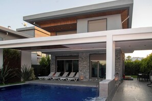Uma casa moderna de dois andares com piscina, cadeiras de descanso e área de varanda.