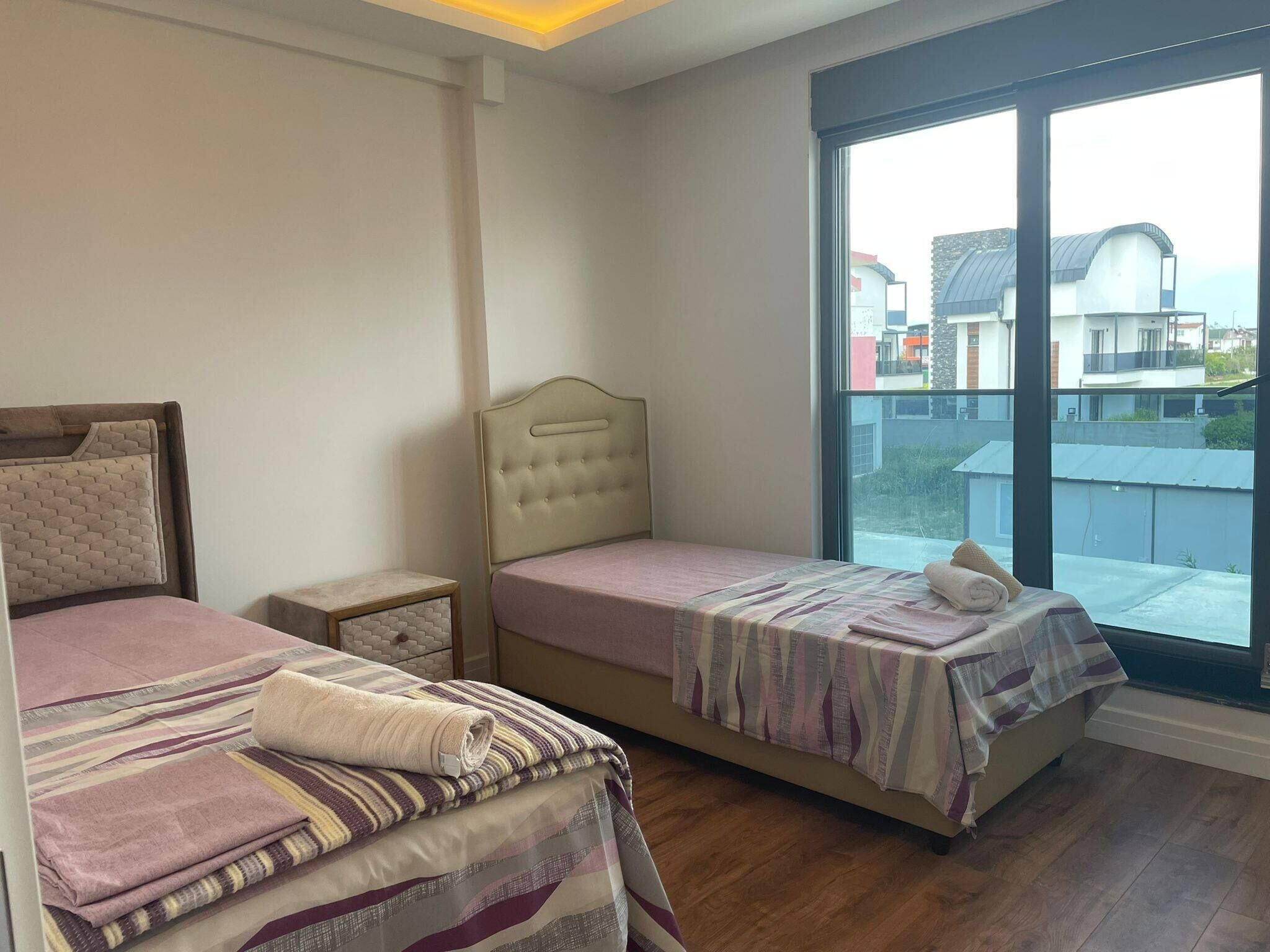 Quarto com duas camas, uma mesa de cabeceira e uma janela grande com vista para edifícios.