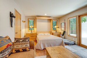 Quarto com cama, mesas de cabeceira, um banquinho de madeira e janela com vista para o verde.