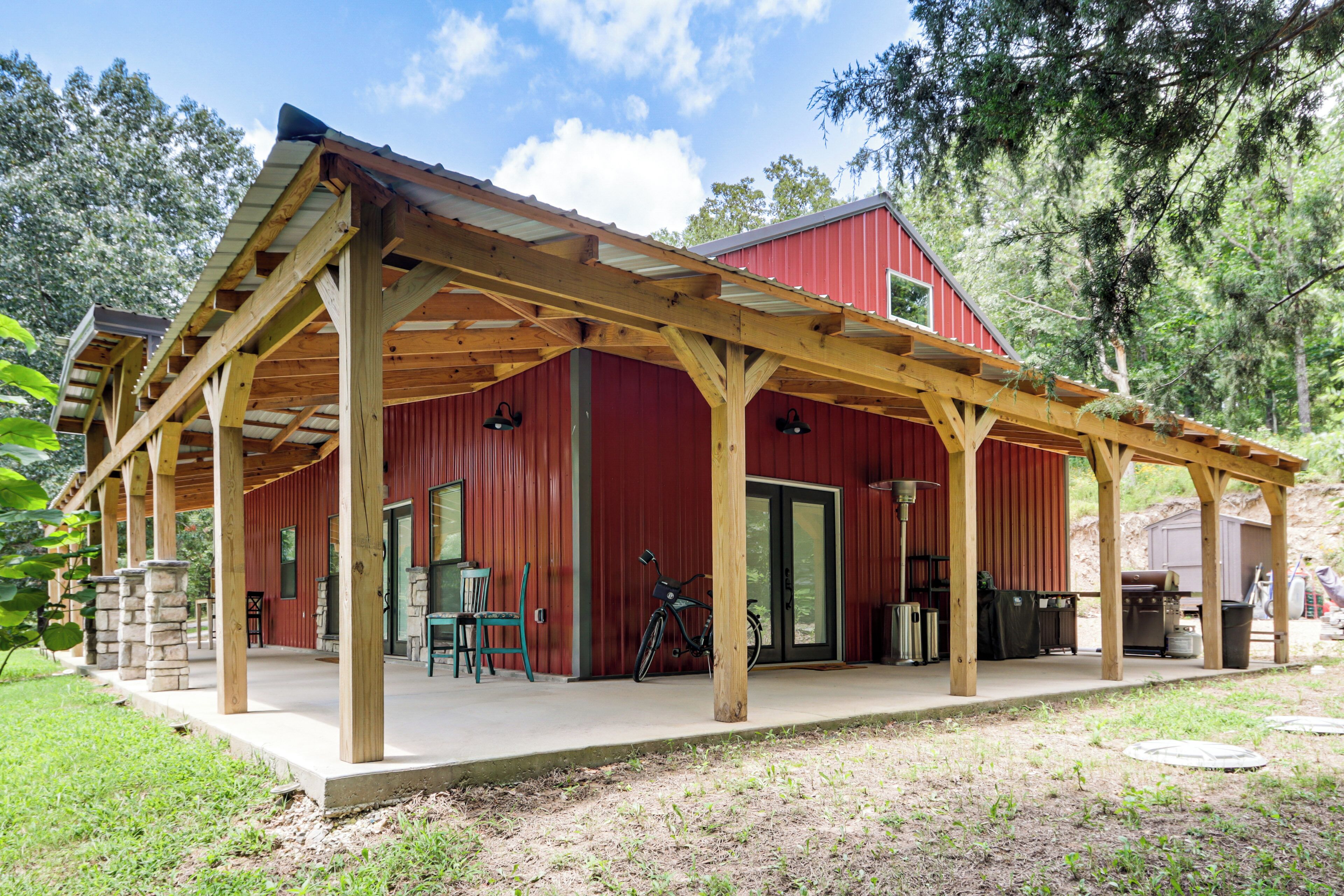 Uma área externa coberta com um edifício vermelho, vigas de madeira, uma mesa, cadeiras e uma bicicleta.