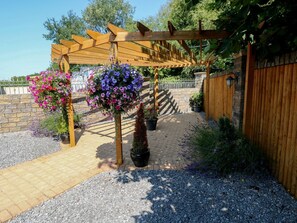Um jardim com pérgola de madeira, cestos de flores suspensos e uma parede de pedra.