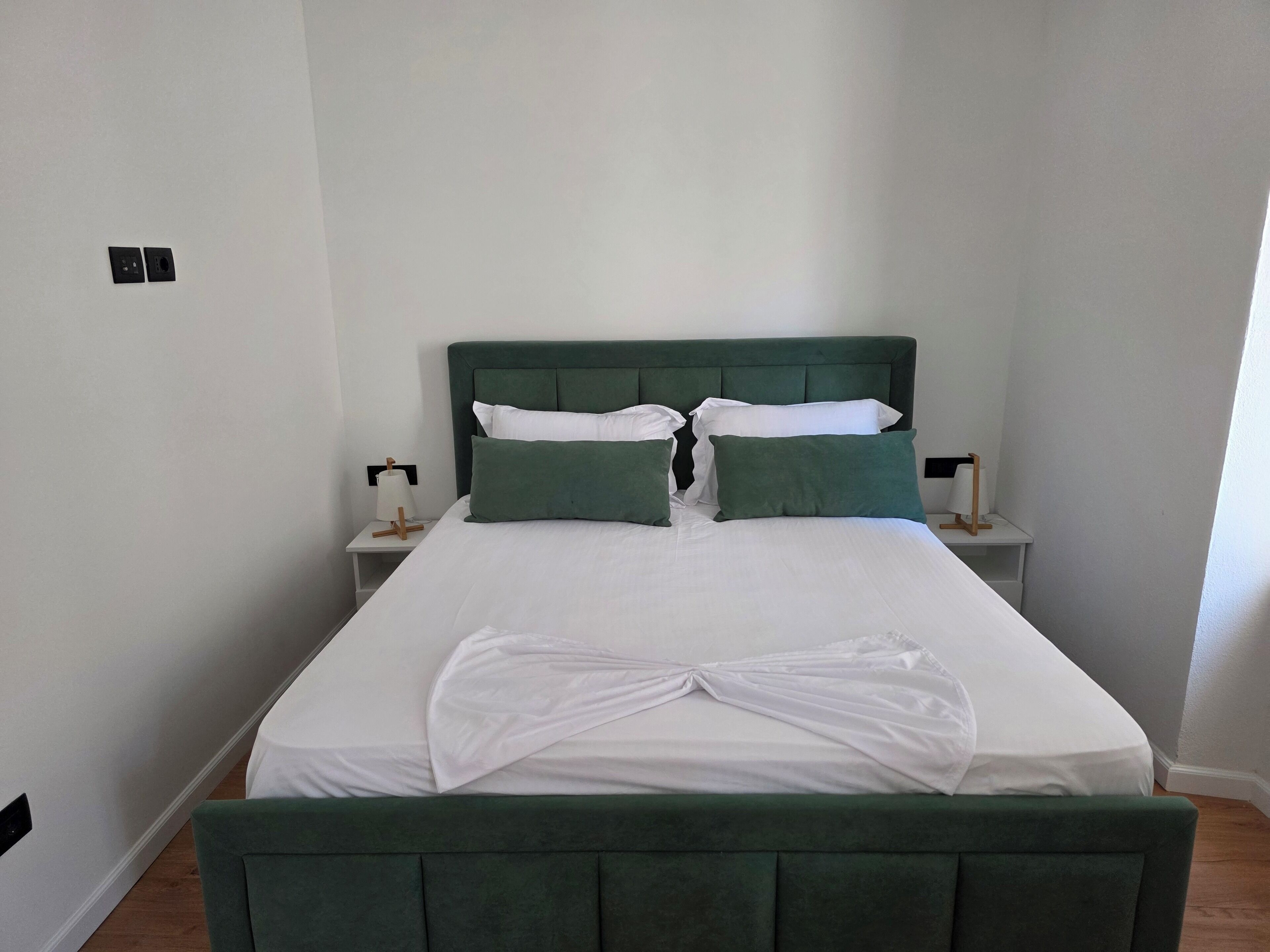 Uma cama bem arrumada com cabeceira verde, roupa de cama branca e duas mesas de cabeceira com abajures.