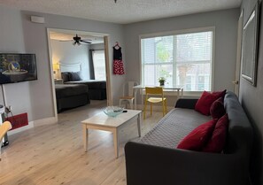 Uma sala de estar com sofá, mesa de centro e mesa de jantar com cadeiras. Há uma televisão fixada na parede e uma janela com persianas.